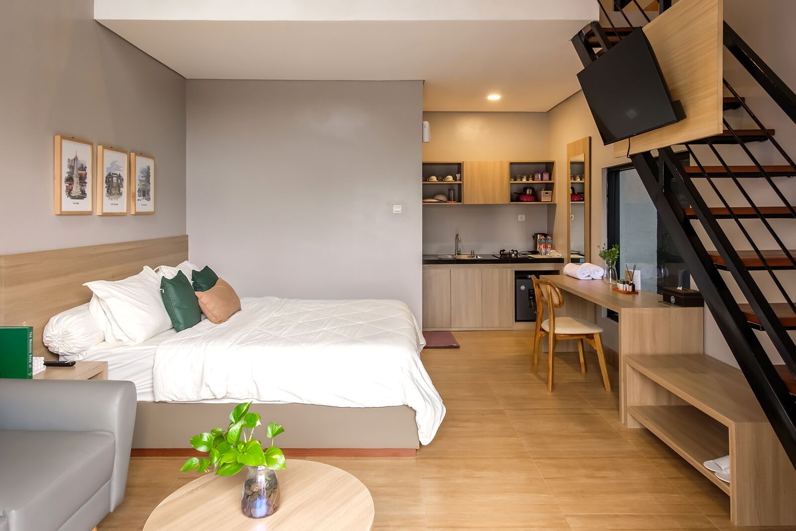 Quarto de hotel compacto com cama, escrivaninha, cadeira, mesinha com planta e cozinha equipada com armários e pia.