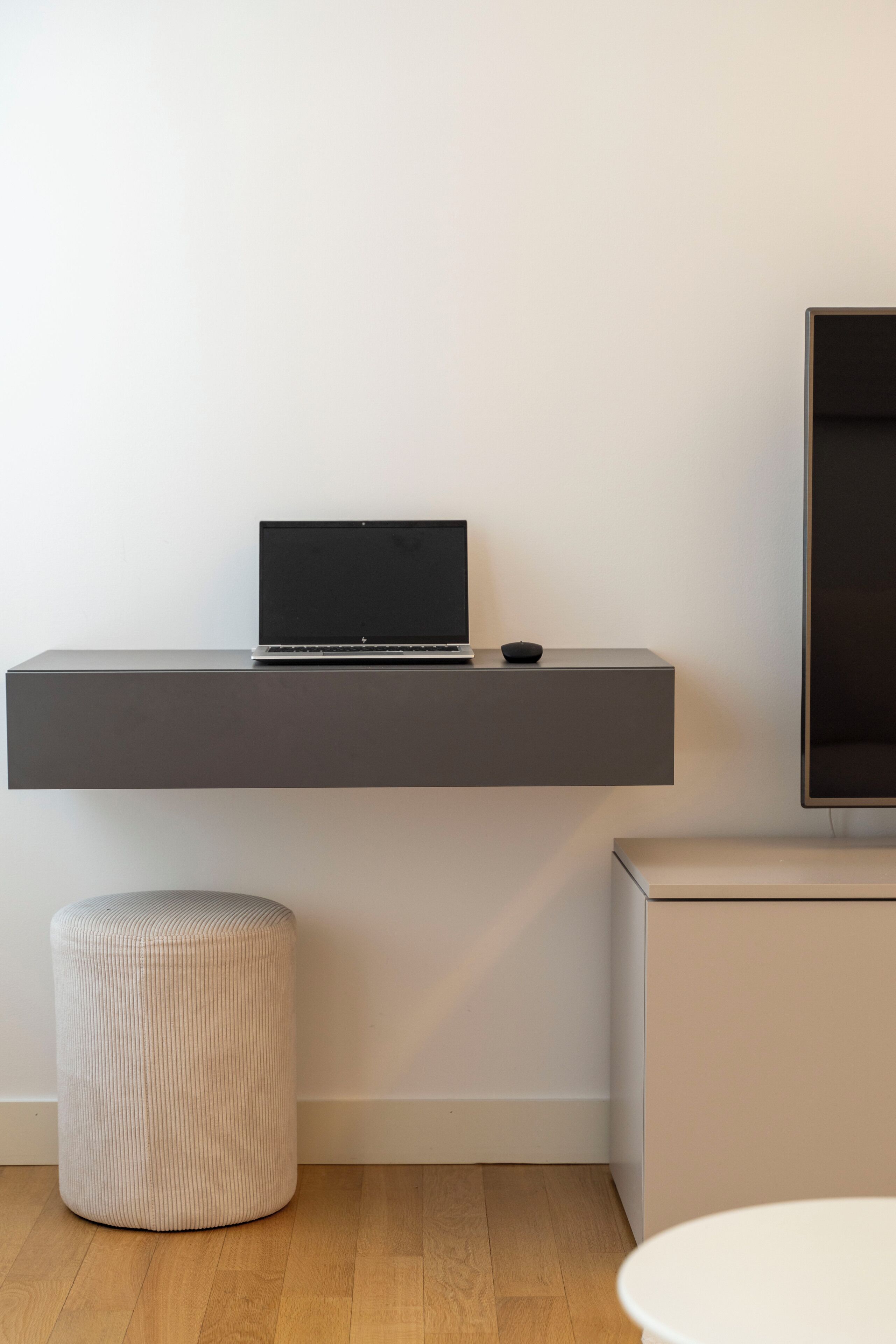 Quarto moderno com um laptop em uma prateleira suspensa, um banquinho redondo e uma mesa branca.