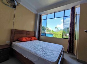 Quarto com cama, mesa de cabeceira, janela com cortinas e ventilador fixado na parede.