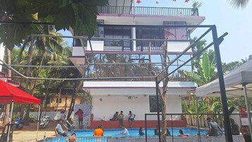 Área de piscina com um edifício de dois andares ao fundo, cercada por palmeiras.