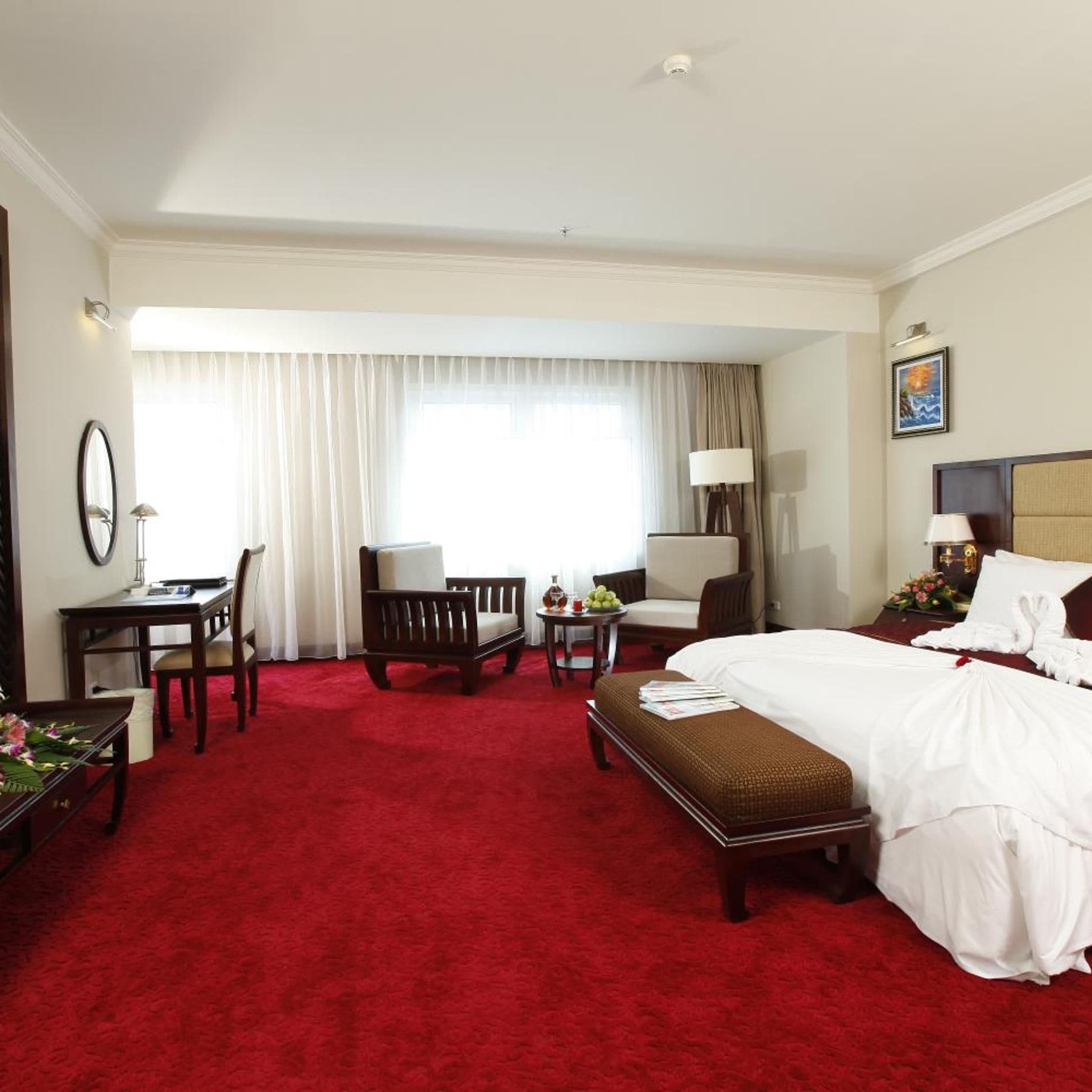 Quarto de hotel com uma cama grande, uma escrivaninha, duas cadeiras, uma mesa pequena e uma janela com cortinas.