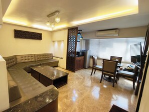 Uma sala moderna com sofá de canto, mesa de centro, área de refeições com cadeiras, televisão e aparelho de ar condicionado.