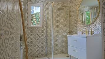 Um banheiro com paredes estampadas, box de vidro, toucador branco com puxadores dourados e espelho redondo.