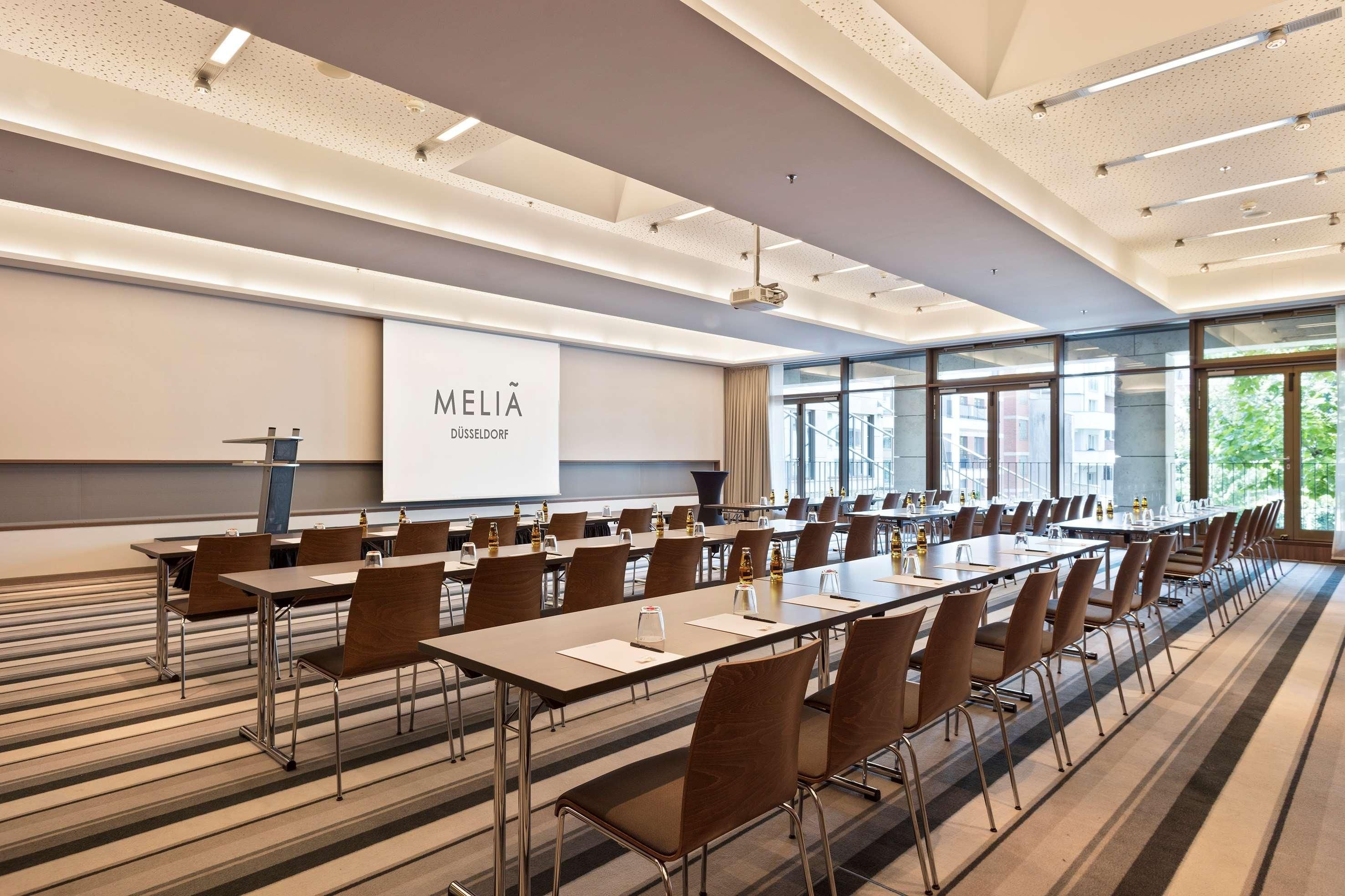 Sala de conferência com uma mesa comprida, cadeiras, tela de projeção e um amplo janelão.