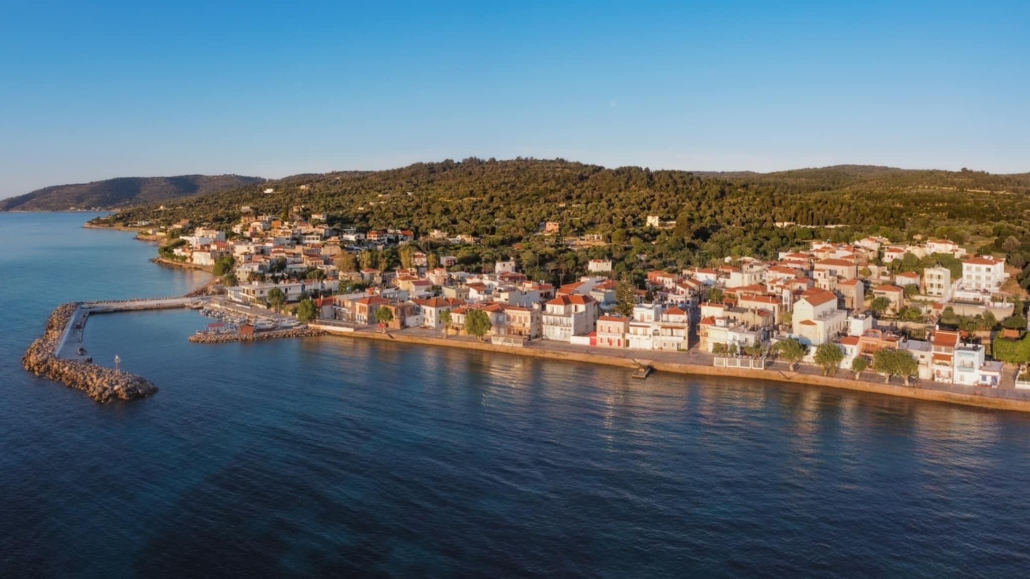 Uma cidade costeira com edifícios ao longo da orla, um molhe rochoso e colinas ao fundo.