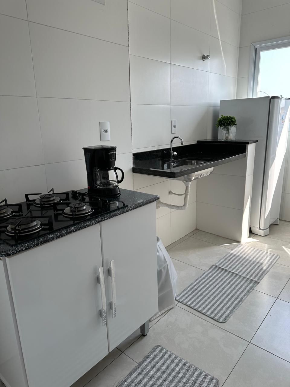 Uma cozinha moderna com armários brancos, bancada preta, cafeteira, fogão a gás, pia de aço inoxidável e uma geladeira.