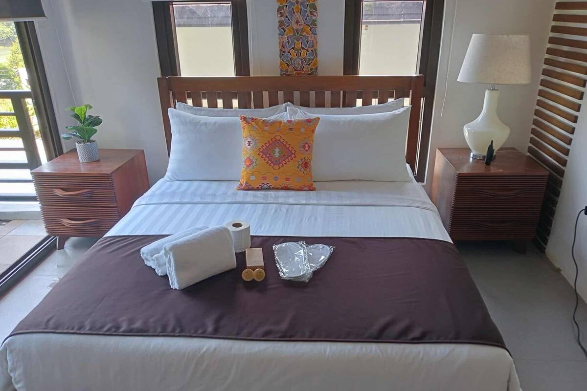 Cama bem arrumada com um cobre-leito marrom, travesseiros brancos e uma almofada decorativa colorida. Há duas mesas de cabeceira de madeira com luminárias, uma planta em vaso e produtos de higiene pessoal na cama.