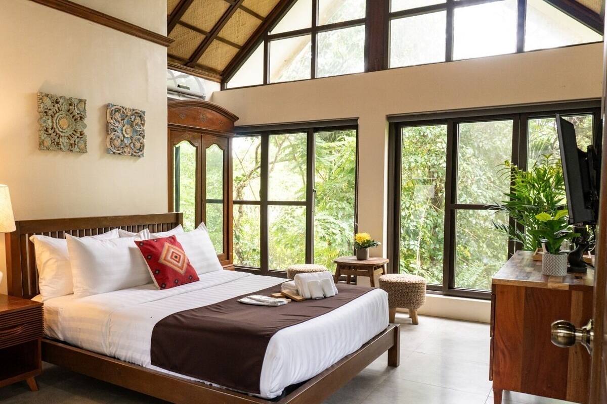 Um quarto espaçoso com uma cama grande, mesa de cabeceira de madeira, escrivaninha e vista para um verde exuberante através de grandes janelas.