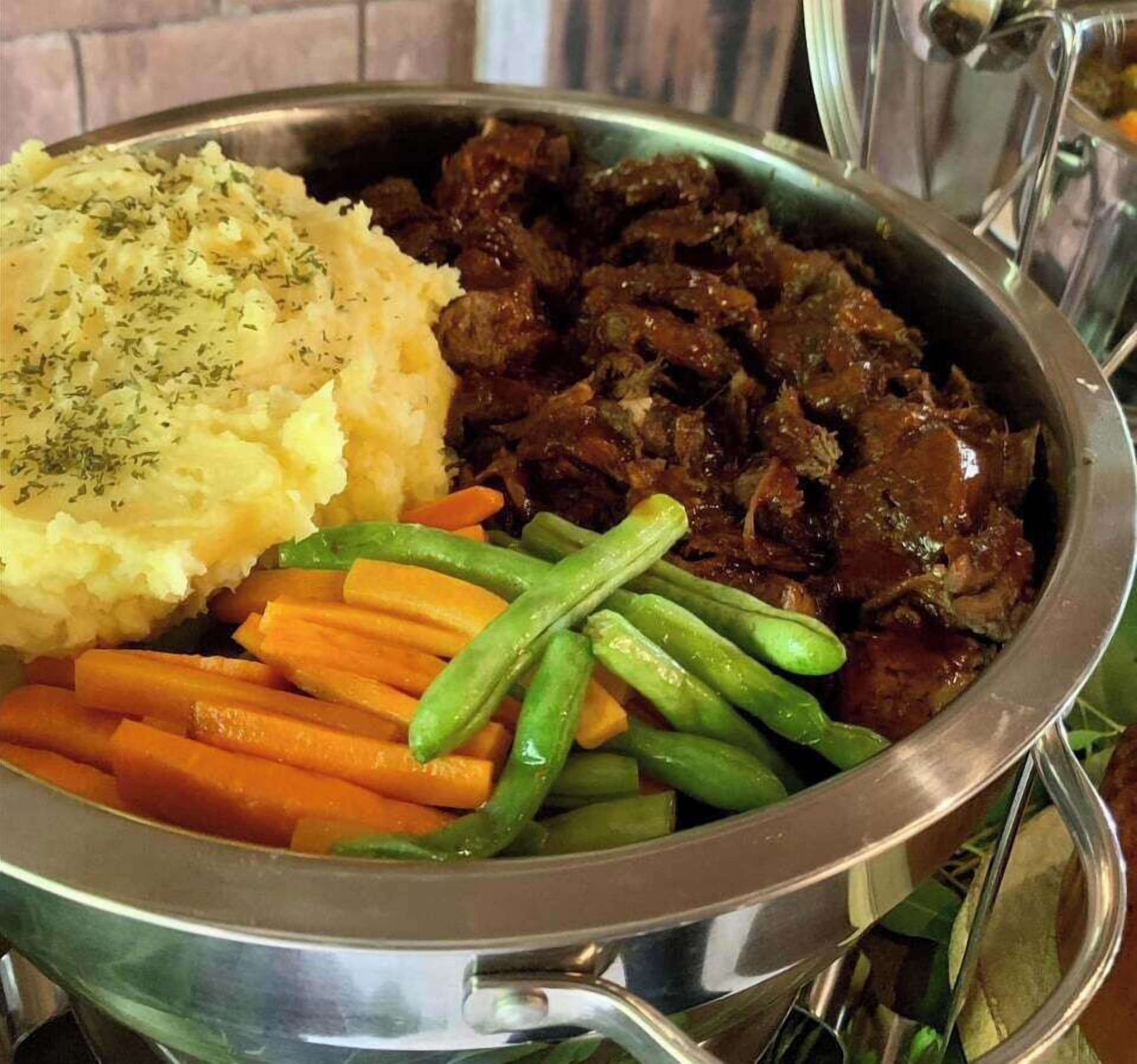 Uma refeição com purê de batata, ensopado de carne, cenouras e vagem, servida em uma tigela de metal.