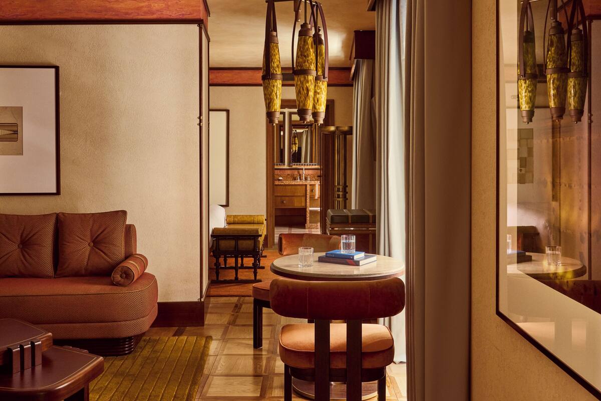 Quarto de hotel moderno com piso de madeira, sofá, mesa de jantar e uma janela grande com cortinas.