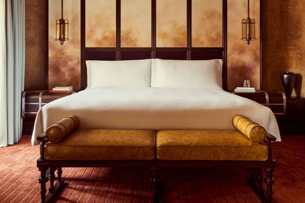 Uma cama grande com colchão branco e duas almofadas, com um banquinho de madeira com travesseiro amarelo. Há duas mesas de cabeceira com abajures, um vaso grande e um livro em uma das mesas.