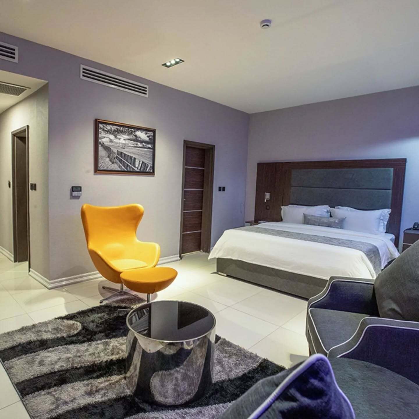 Quarto de hotel moderno com uma cama grande, uma poltrona amarela vibrante, uma pequena mesa redonda e um quadro na parede.