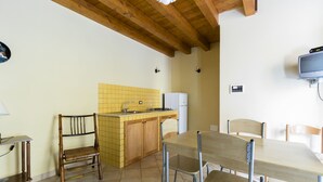 Uma cozinha compacta com teto de madeira, painel de azulejos e uma pequena geladeira. Há uma mesa de jantar com cadeiras e uma cadeira de madeira.