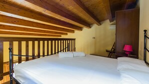 Quarto com teto de madeira, cama com lençóis brancos, abajur vermelho em uma mesa de cabeceira e uma escada encostada na parede.