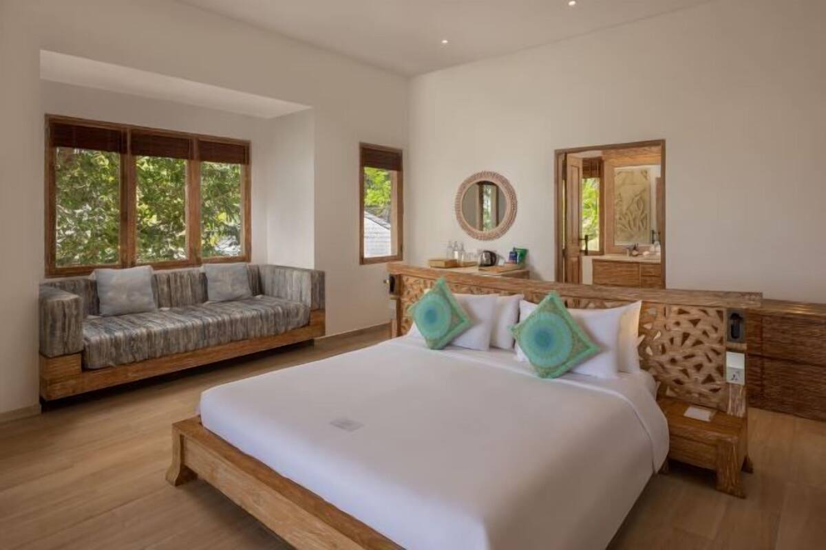 Quarto com cama, sofá, cabeceira de madeira, espelho e janela com vista para árvores.