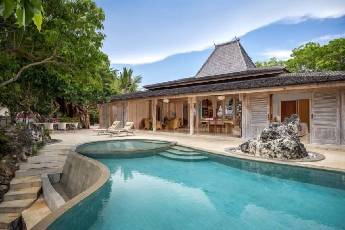Piscina com escadas de acesso, área externa coberta com mobiliário e um edifício tradicional com telhado de azulejos.