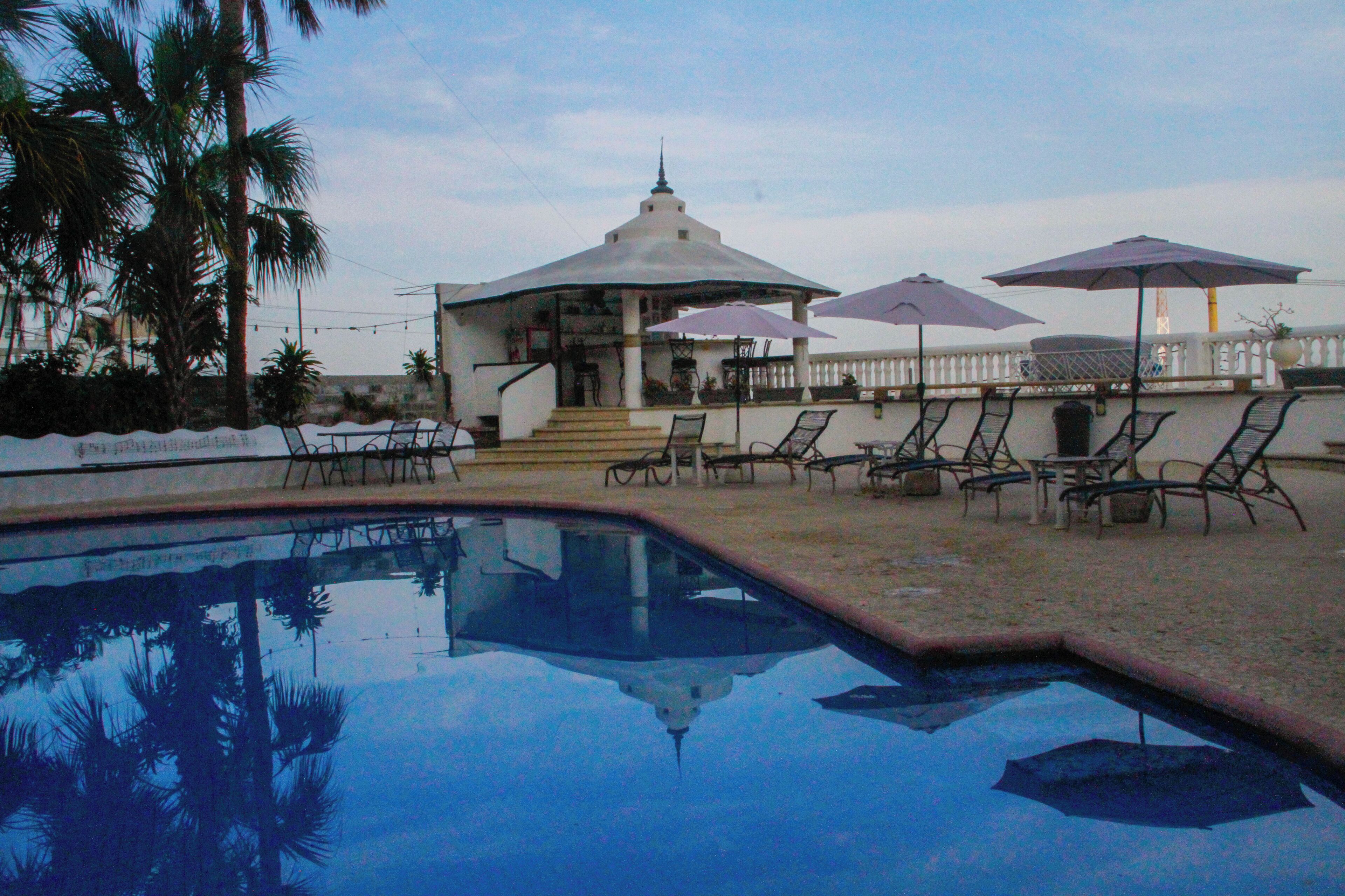 Uma área de piscina com espreguiçadeiras e guarda-sóis, um gazebo e palmeiras.