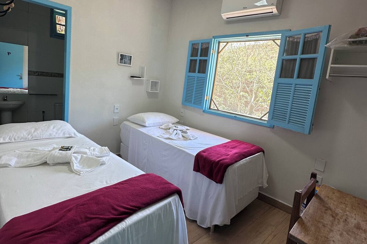 Quarto de hotel com duas camas, uma mesa de madeira, uma janela com persianas azuis e um aparelho de ar condicionado.