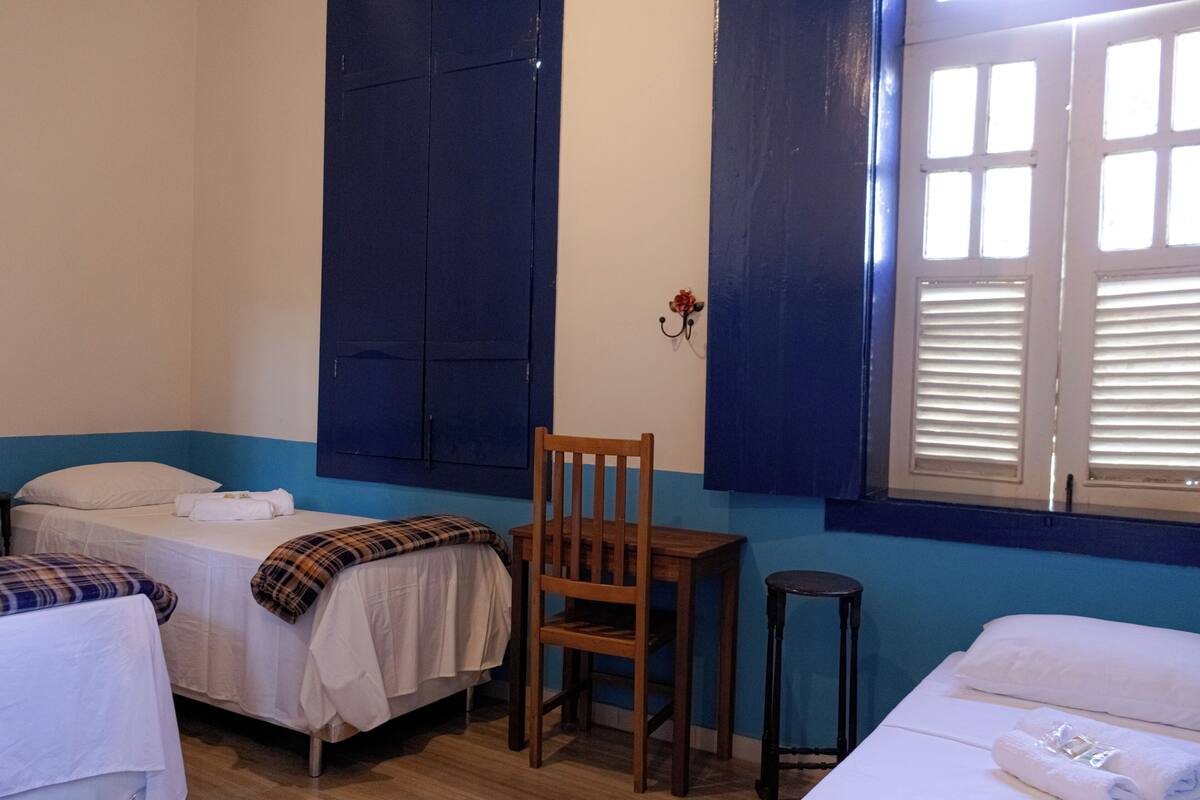 Quarto com duas camas, uma cadeira de madeira, uma mesa pequena e uma janela com persianas.
