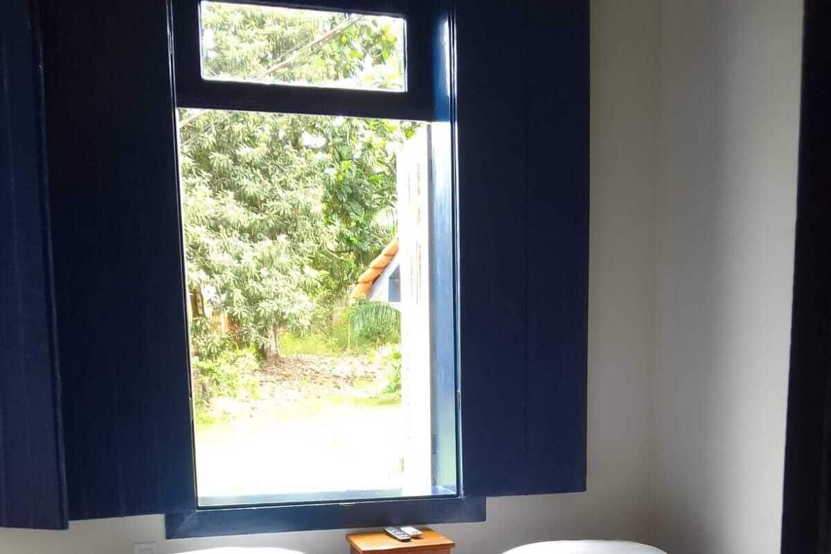 Quarto com duas camas, janela com cortinas, uma pequena mesa de madeira e vista para o verde lá fora.