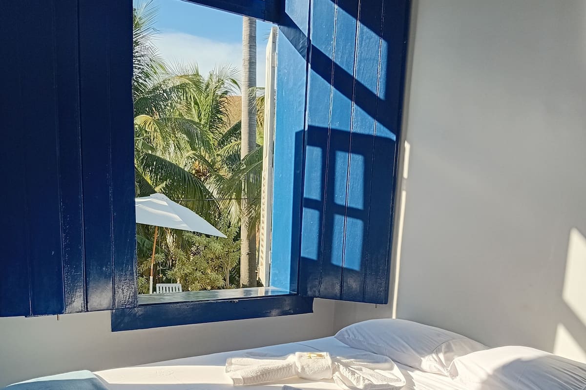 Quarto com cama, janela com persianas azuis e vista para palmeiras lá fora.