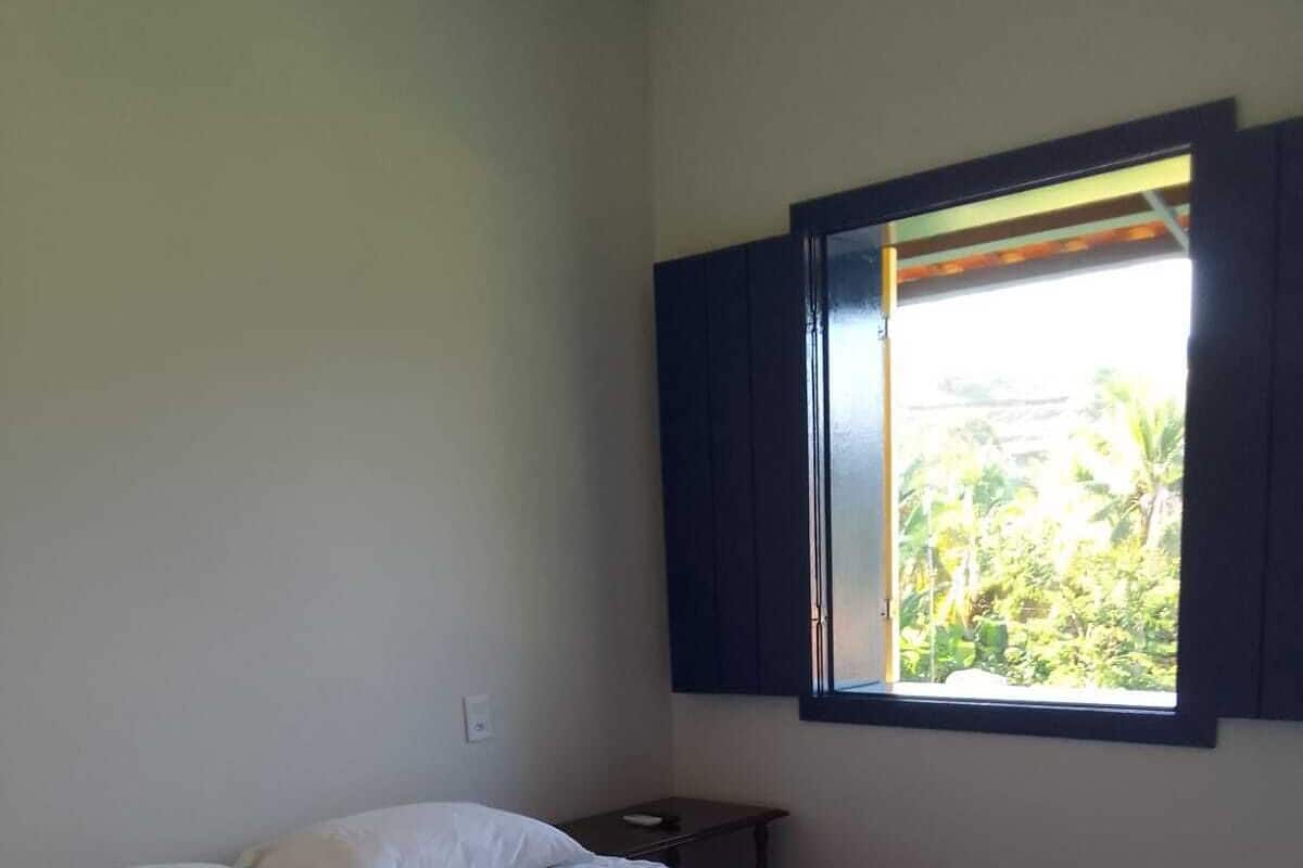 Cama bem arrumada com lençóis brancos e manta bege, mesa de cabeceira com controle remoto e janela com persianas escuras dando para o verde.