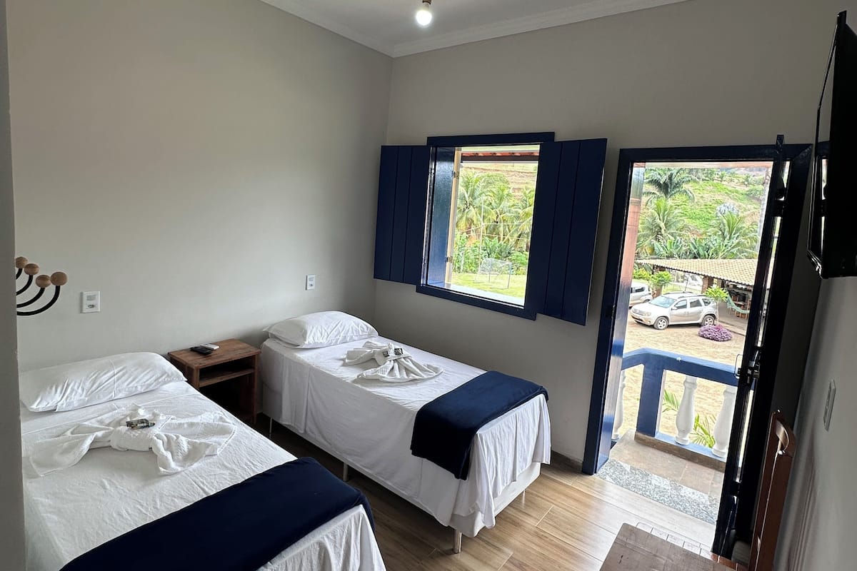Quarto de hotel com duas camas de solteiro, uma mesa de madeira e uma janela com persianas azuis.