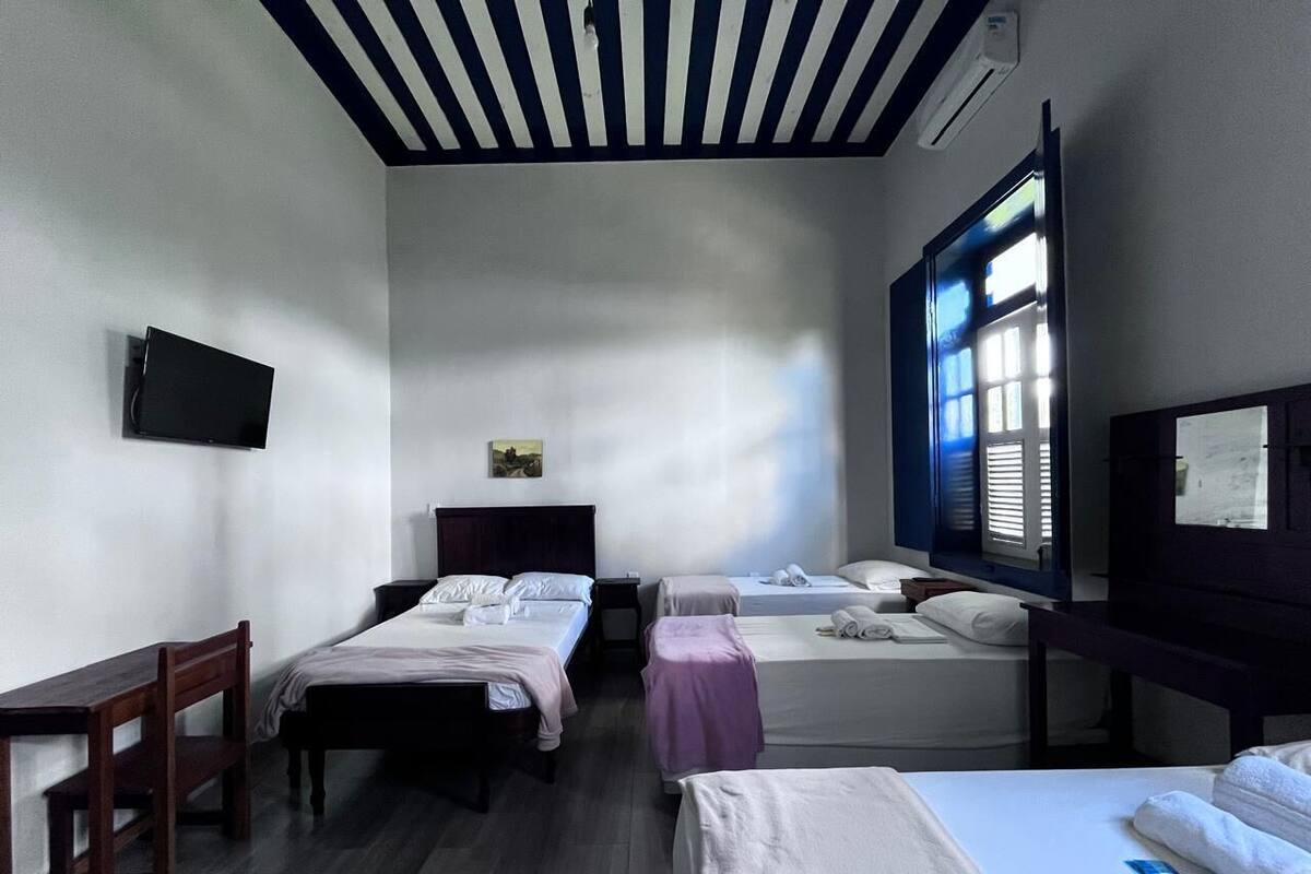 Quarto de hotel com duas camas, uma mesa, uma cadeira, uma TV e uma janela com persianas.