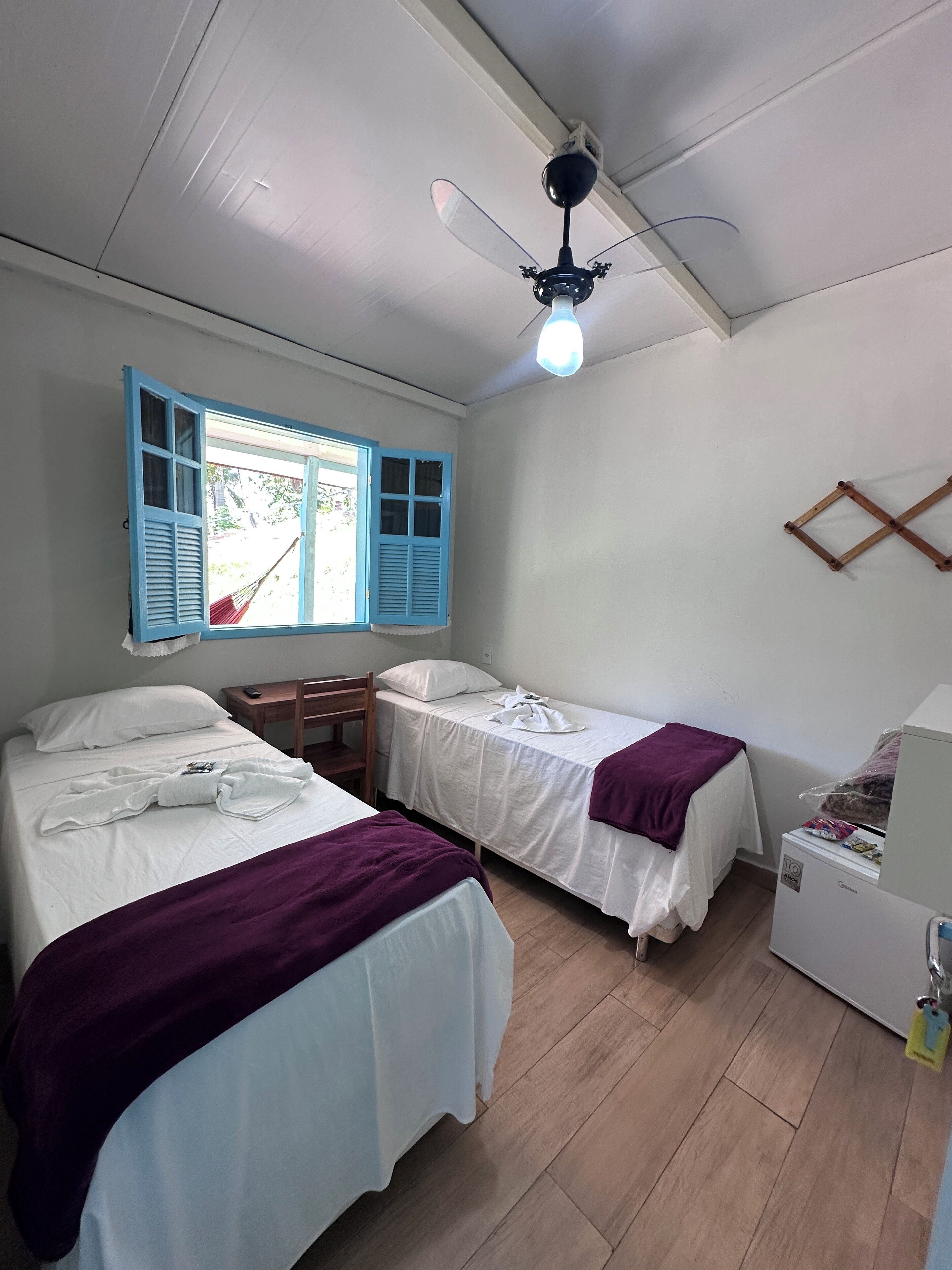 Quarto com duas camas, ventoinha no teto, janela com persianas azuis e um pequeno frigorífico.