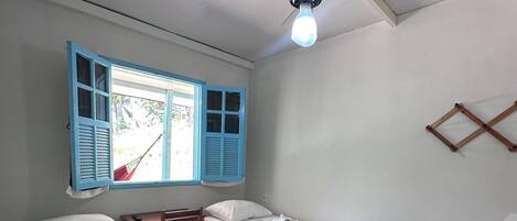 Quarto com duas camas, ventoinha no teto, janela com persianas azuis e um pequeno frigorífico.