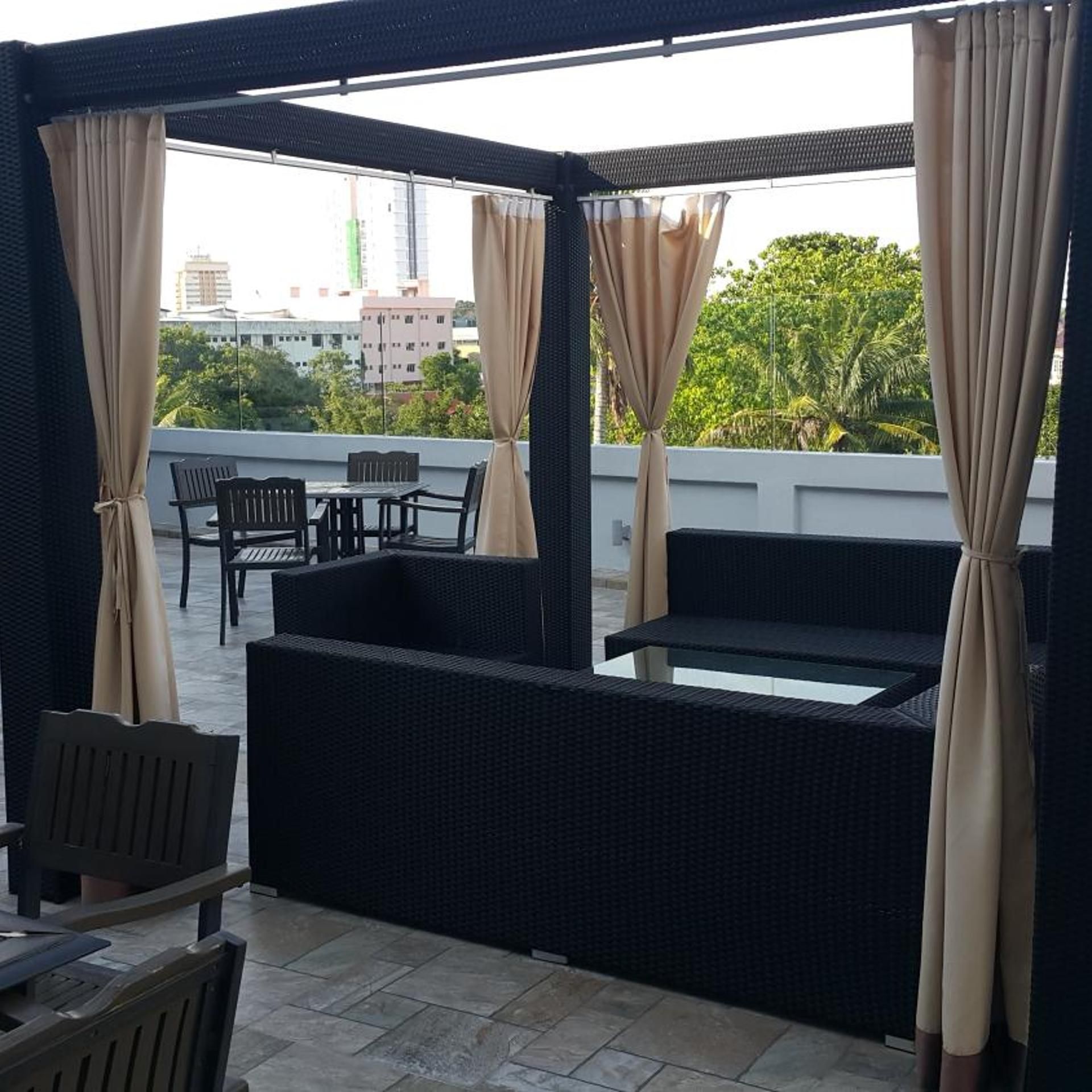 Área de estar externa com mesa e cadeiras, cercada por móveis de vime escuro e cortinas bege.