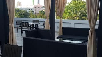 Área de estar externa com mesa e cadeiras, cercada por móveis de vime escuro e cortinas bege.