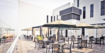 Uma área de terraço ao ar livre com várias mesas e cadeiras, guarda-sóis e um edifício moderno ao fundo.