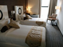 Quarto de hotel com duas camas, mesa de cabeceira, uma cadeira e uma janela grande.