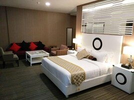 Quarto de hotel com cama, área de estar com sofá e poltronas, mesa pequena e mesa de cabeceira com abajur.