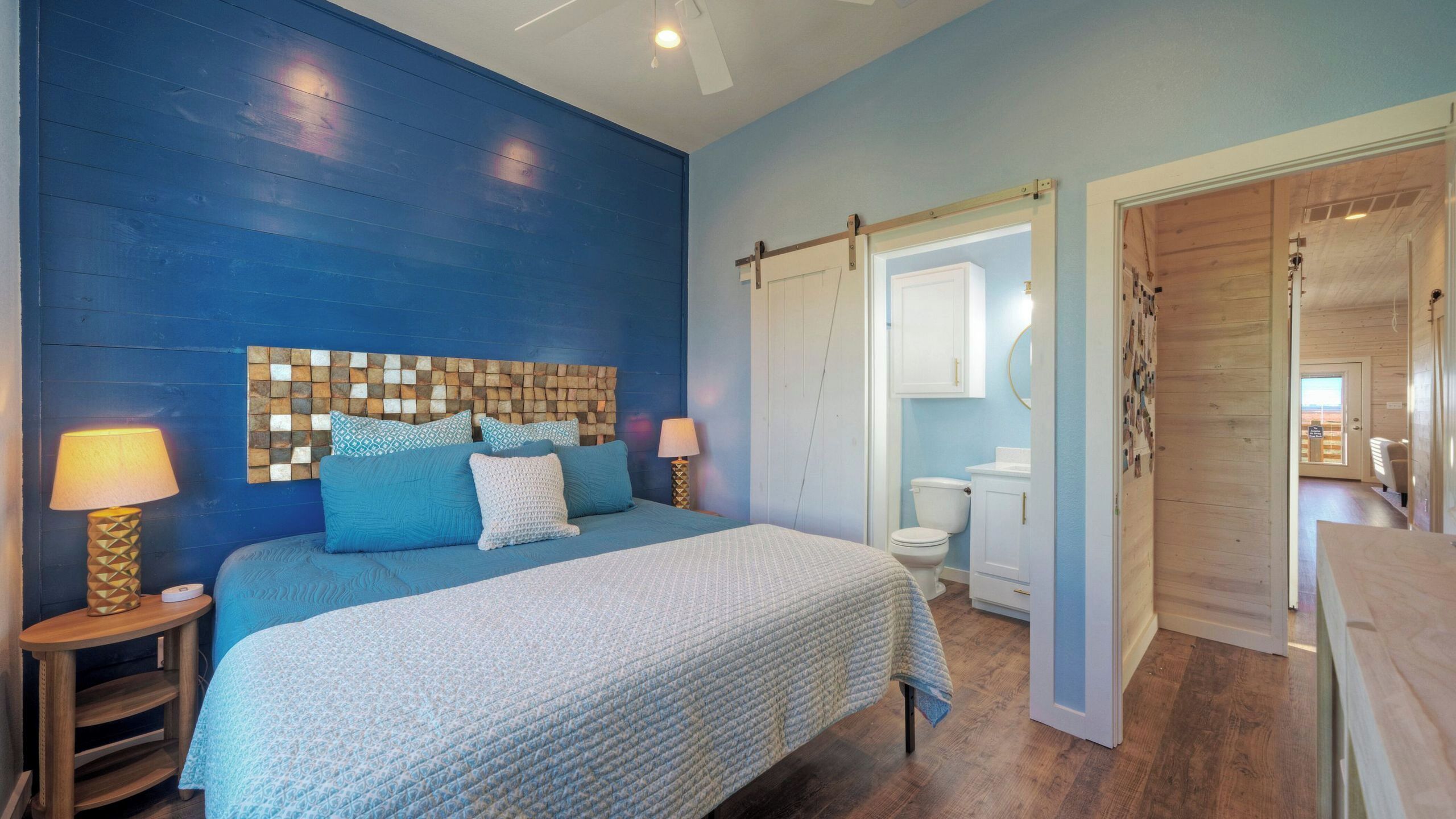 Quarto com parede azul, cabeceira de madeira, cama com roupa de cama azul, duas luminárias de mesa e banheiro com vaso sanitário branco. Porta de madeira leva a outro cômodo.