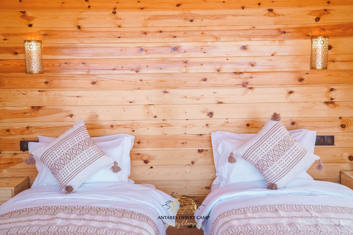 Interior de uma cabana de madeira com duas camas, almofadas estampadas e luminárias fixadas na parede.