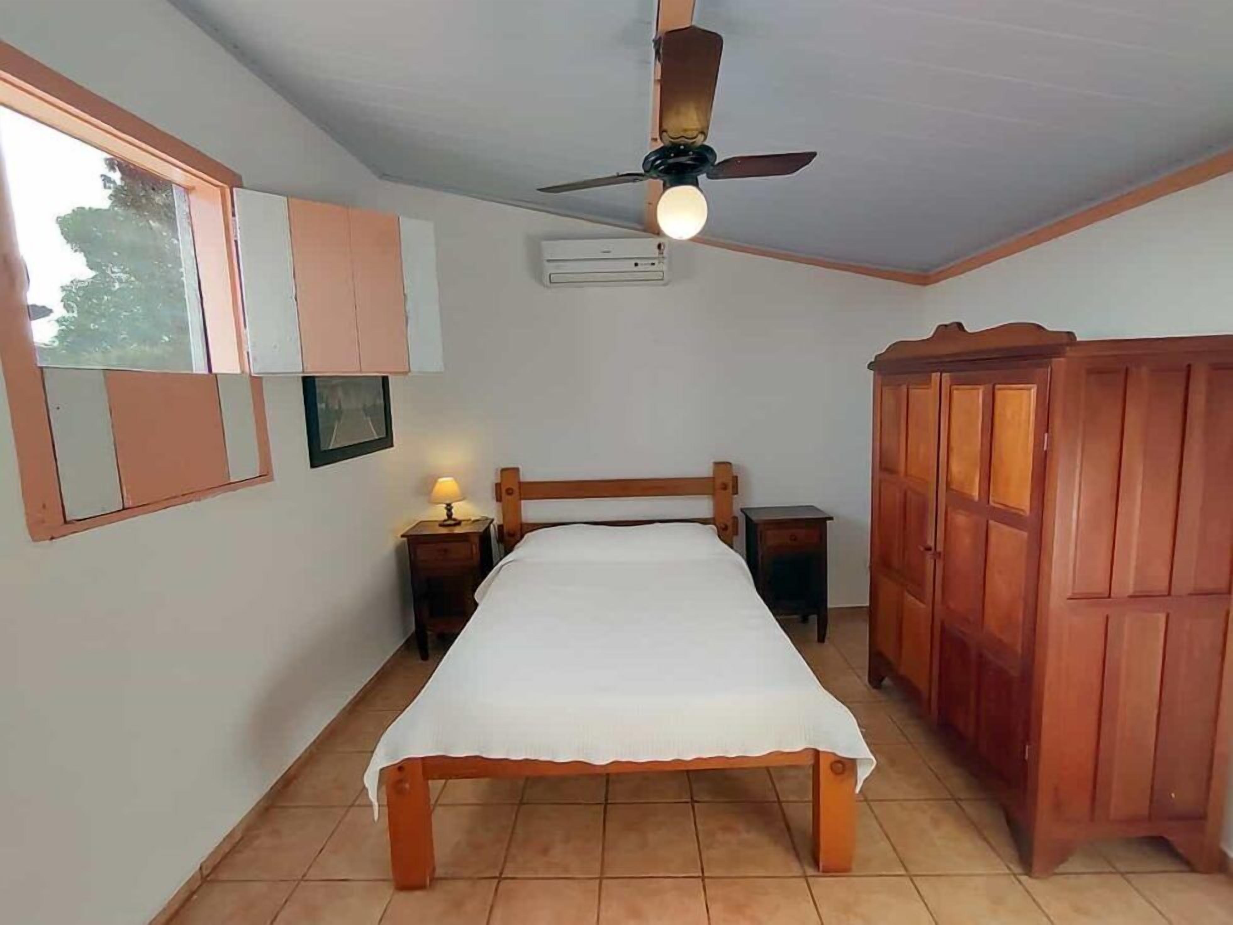Um quarto com cama, mesa de cabeceira, guarda-roupa de madeira, ventoinha no teto e janela.
