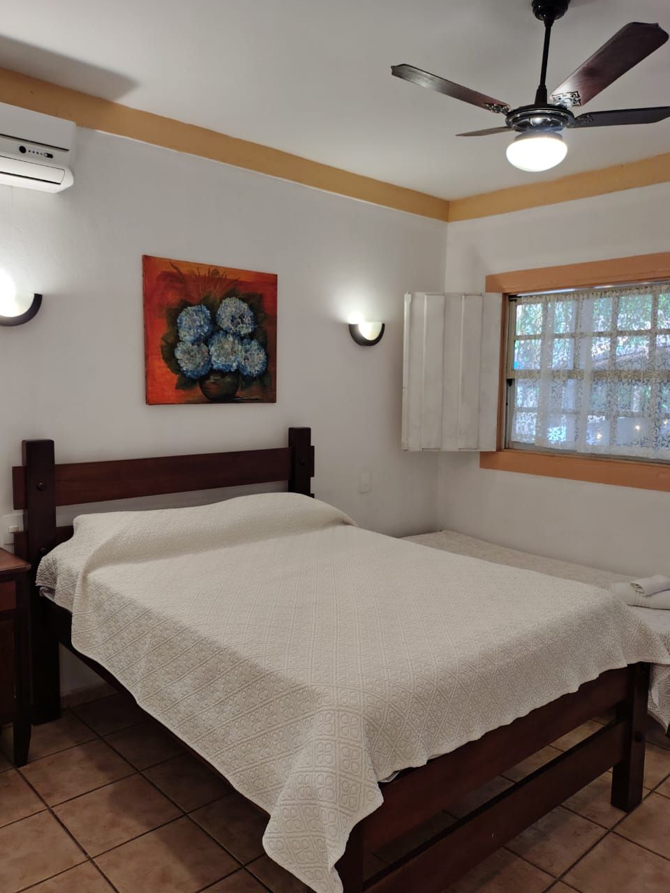 Uma cama de solteiro com colcha branca num quarto com ventoinha no teto, ar condicionado fixado na parede, pintura de flores e janela com vista.