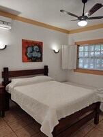 Uma cama de solteiro com colcha branca num quarto com ventoinha no teto, ar condicionado fixado na parede, pintura de flores e janela com vista.