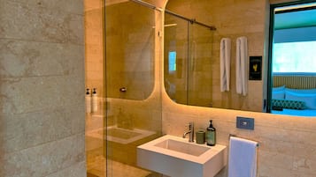 Um banheiro moderno com chuveiro em vidro, pia branca e um espelho grande refletindo uma cama com travesseiros.