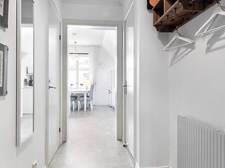 Um corredor com paredes brancas, uma prateleira de madeira com cabides, um radiador e uma porta que dá para uma sala de jantar.