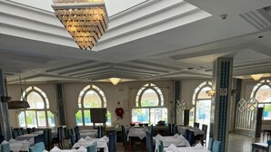 Uma ampla sala de jantar com várias mesas e cadeiras, grandes janelas em arco e um candelabro.