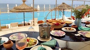 Mesa de jantar com diversos pratos e bebidas, com vista para uma piscina e o oceano.