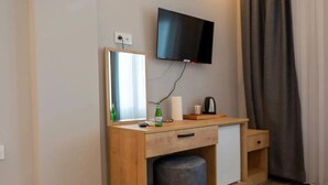 Quarto compacto com uma escrivaninha de madeira, um espelho, uma televisão fixada na parede e um banquinho pequeno.