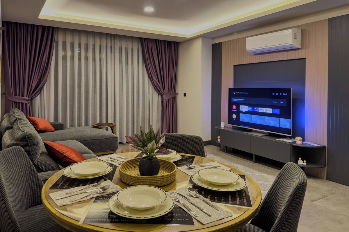 Uma sala de estar moderna com área de jantar, apresentando uma TV de tela plana, um sofá e uma mesa de jantar posta com pratos e talheres.