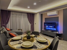 Uma sala de estar moderna com área de jantar, apresentando uma TV de tela plana, um sofá e uma mesa de jantar posta com pratos e talheres.