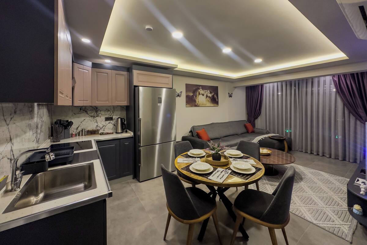 Uma cozinha moderna com geladeira embutida, uma sala de jantar com mesa redonda e quatro cadeiras, e uma sala de estar com sofá e cortinas.