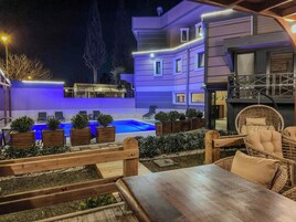 Uma área moderna de piscina ao ar livre com espreguiçadeiras de madeira e vasos de plantas, rodeada por um edifício bem iluminado com varanda.