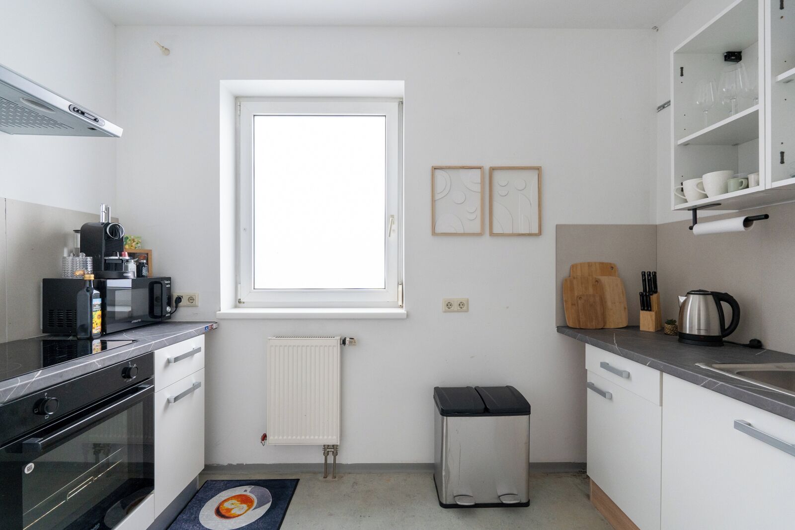 Cozinha moderna com forno embutido, micro-ondas e máquina de café. Há uma janela, um radiador e uma lixeira.